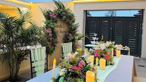 Montagem para um evento ao ar livre com uma mesa decorada com arranjos florais e velas, uma placa com a inscrição "Muweta's Bridal Shower" e um painel de folhagem e flores.