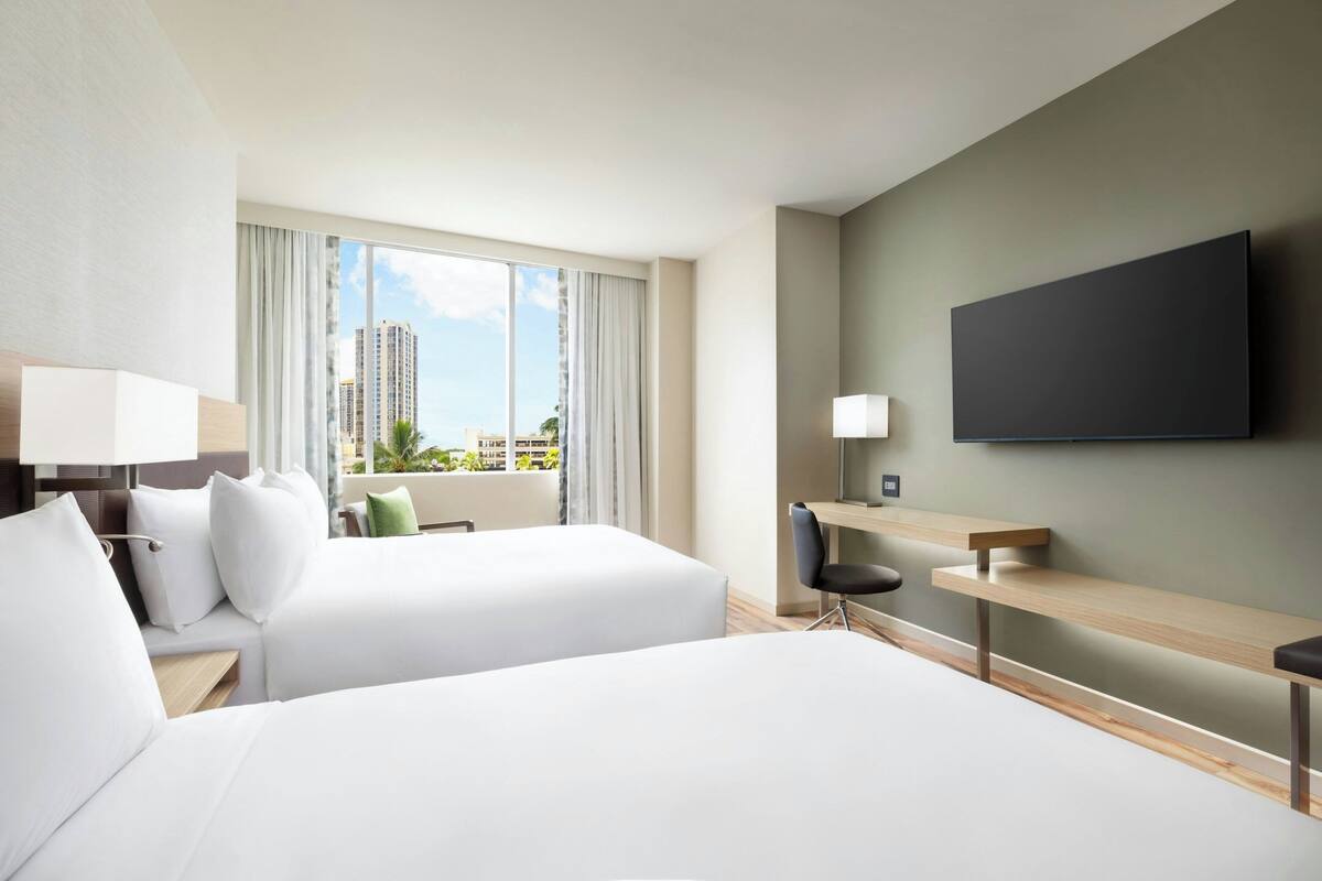 Quarto de hotel com duas camas, uma escrivaninha, uma cadeira, televisão de tela plana e uma janela grande com cortinas.