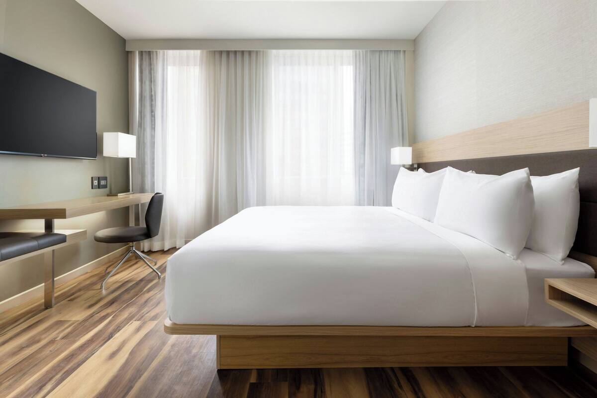 Quarto de hotel com uma cama grande, uma escrivaninha com cadeira, televisão de tela plana e uma janela ampla com cortinas.