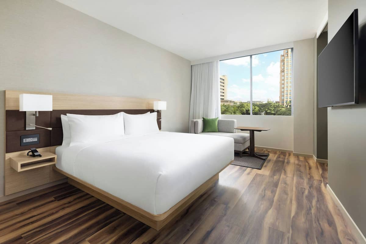 Quarto de hotel com uma cama grande, mesa de cabeceira com luminária, pequena mesa com cadeira e televisão na parede.