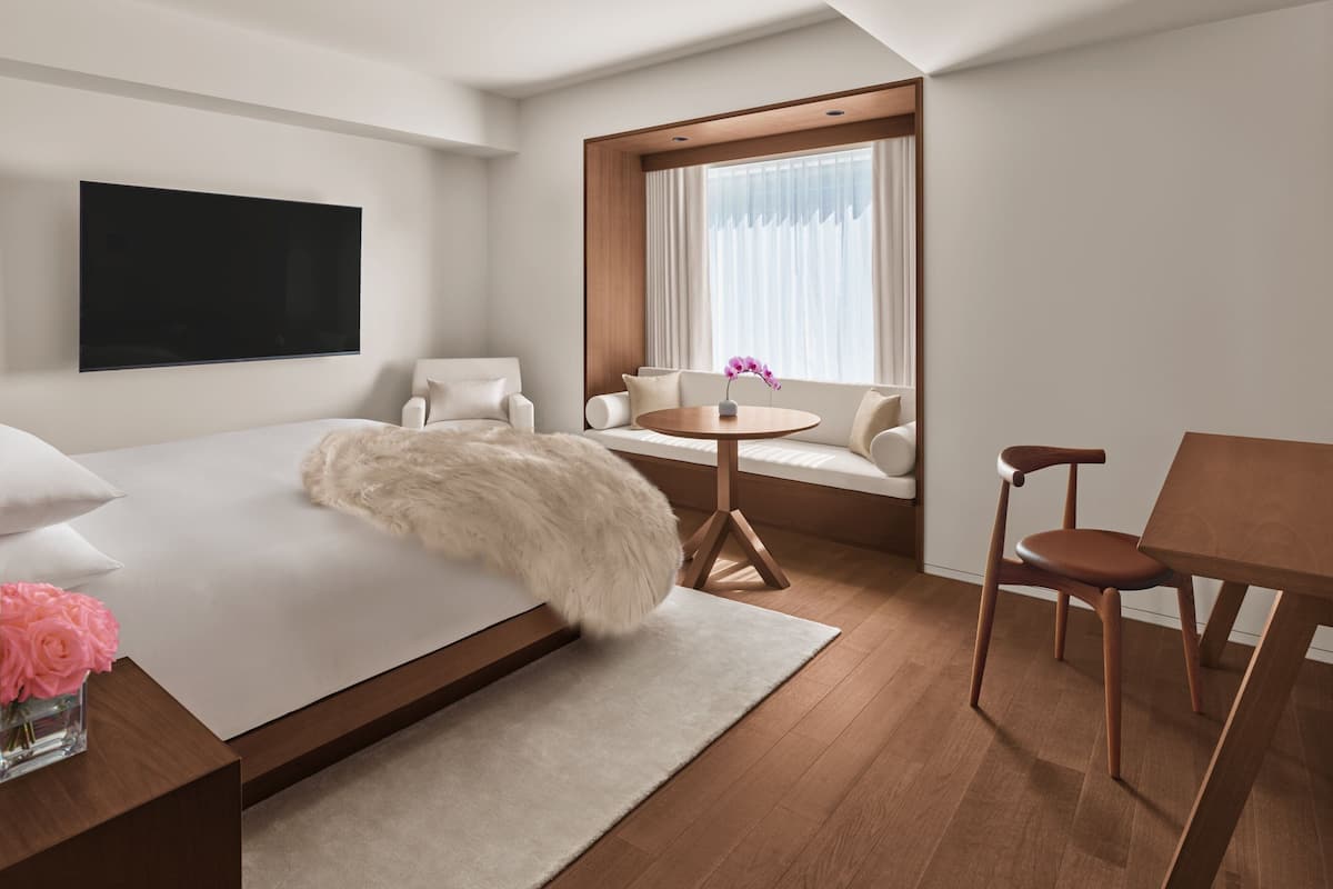 Quarto de hotel moderno com uma cama grande, televisão na parede, uma mesinha redonda com uma cadeira e uma janela com cortinas.
