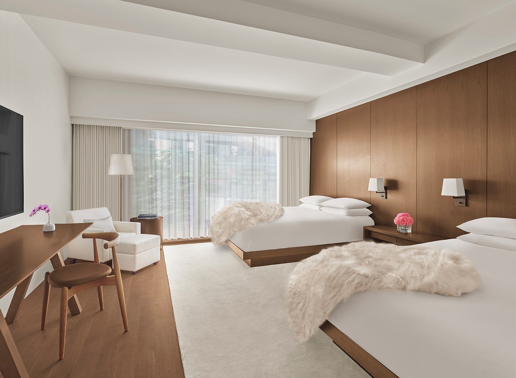 Quarto de hotel moderno com uma cama grande, uma escrivaninha com cadeira, uma mesinha com um vaso de flores e uma janela ampla com cortinas.