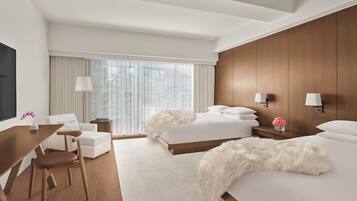 Quarto de hotel moderno com uma cama grande, uma escrivaninha com cadeira, uma mesinha com um vaso de flores e uma janela ampla com cortinas.