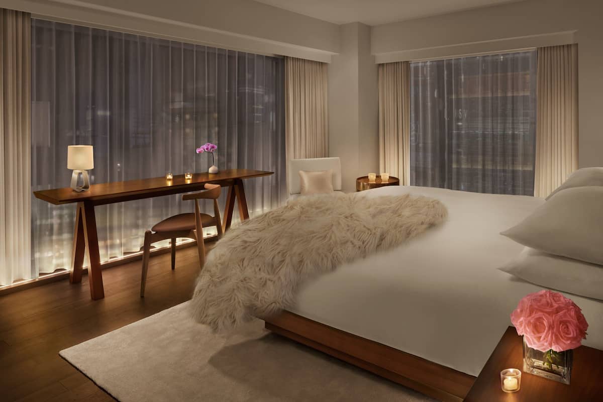 Quarto de hotel com uma cama grande, uma escrivaninha, uma cadeira e uma mesa de cabeceira com um vaso de flores.