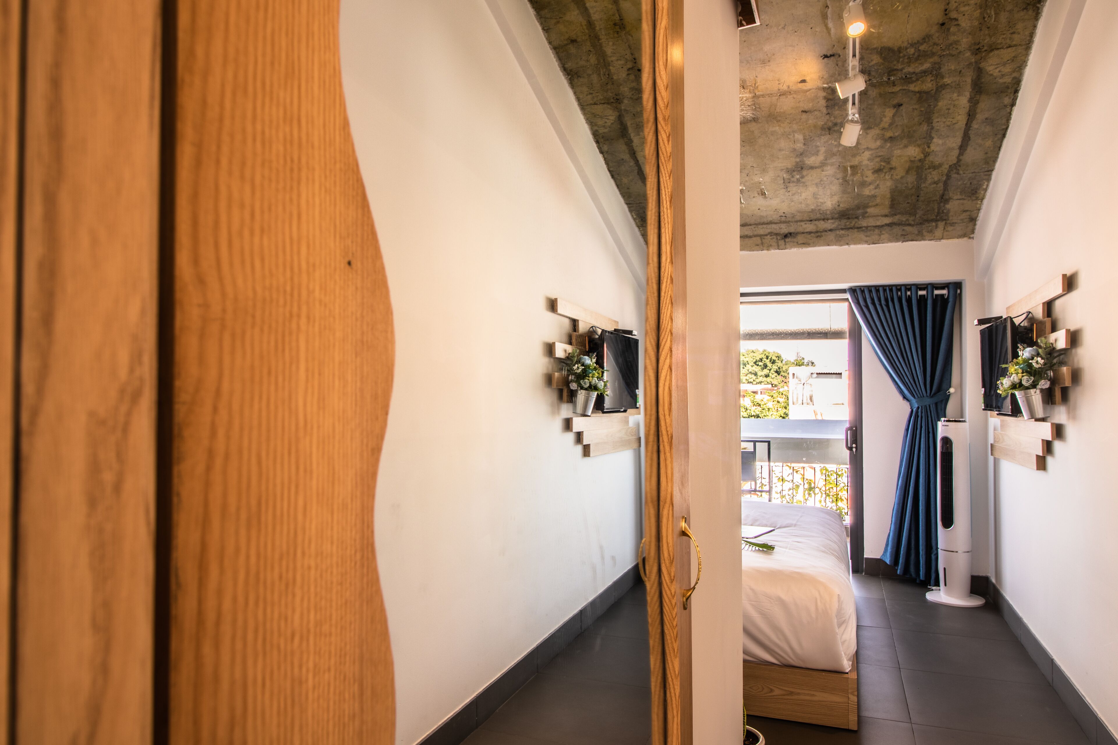 Corredor estreito que dá para um quarto, com cama, mesa de cabeceira e janela com cortinas.