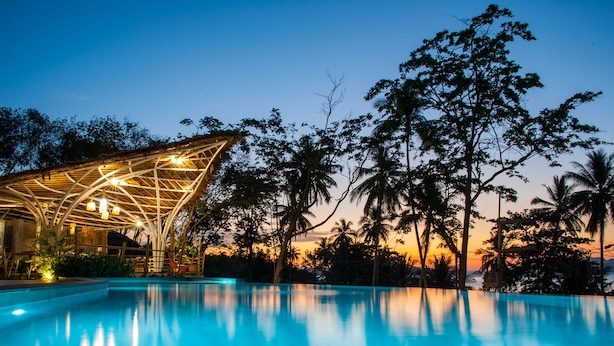 Uma área ao lado da piscina com uma moderna estrutura de gazebo, rodeada por palmeiras e um céu claro.