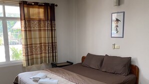 Uma cama bem arrumada, com estrutura de madeira e edredom bege e castanho, uma mesa de cabeceira com abajur e uma janela com cortinas.
