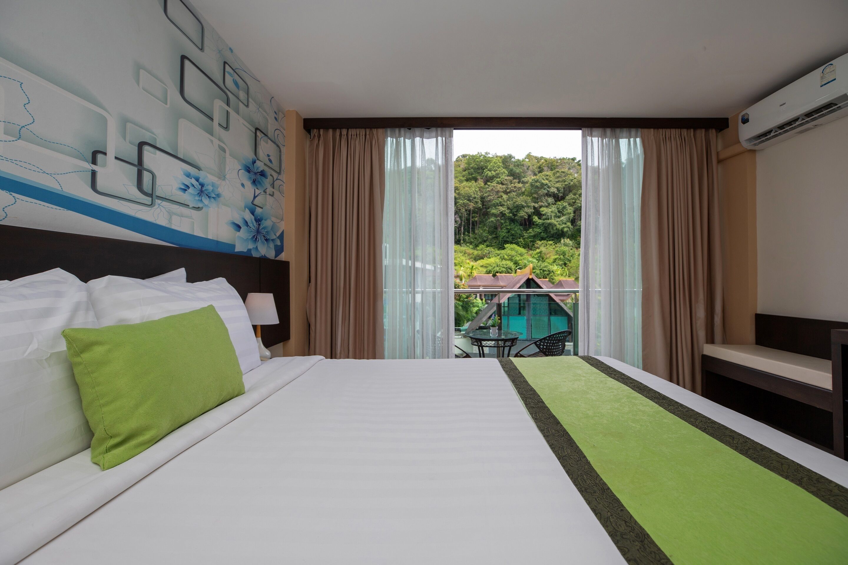Quarto de hotel com uma cama grande, um travesseiro verde, vista para uma varanda com mesa e cadeiras, e um quadro na parede.
