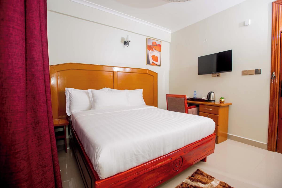 Quarto de hotel com cama de madeira, televisão fixada na parede, escrivaninha com cadeira e cortina vermelha.