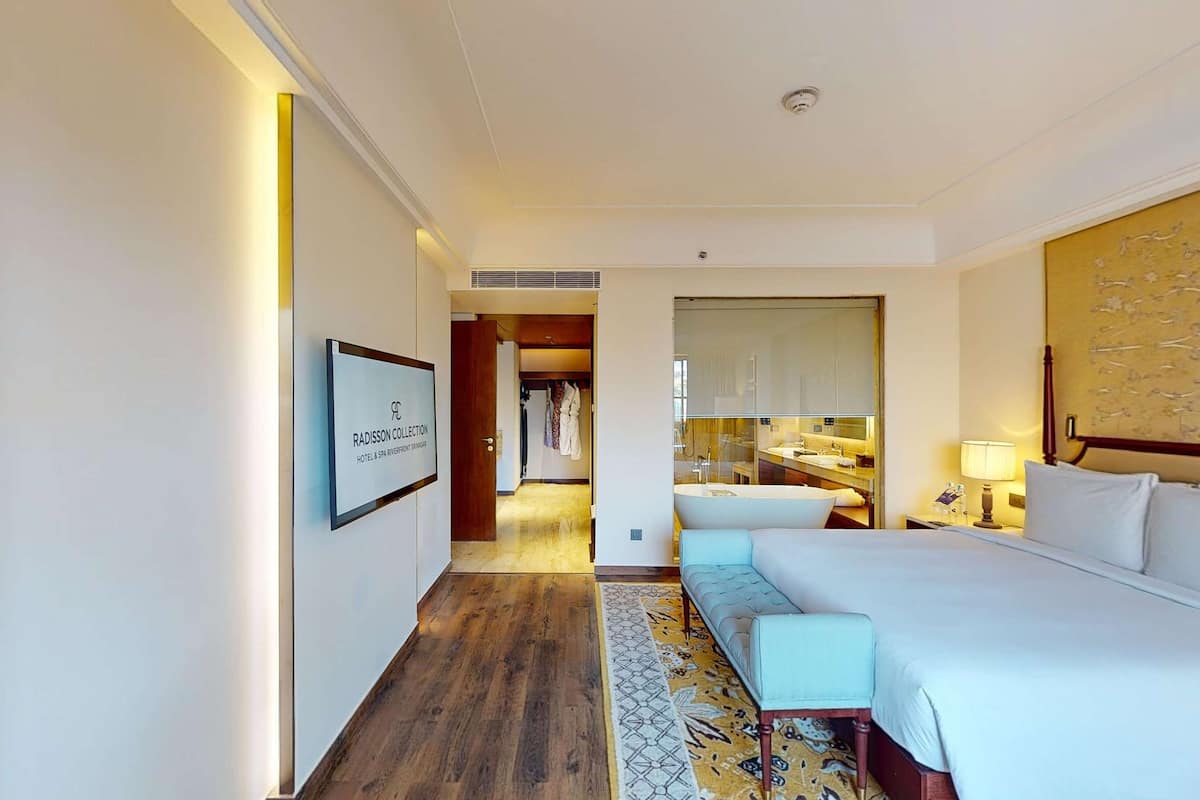 Quarto de hotel com uma cama grande, uma cama menor, área de estar, banheiro com um espelho grande e corredor que dá para outro quarto.