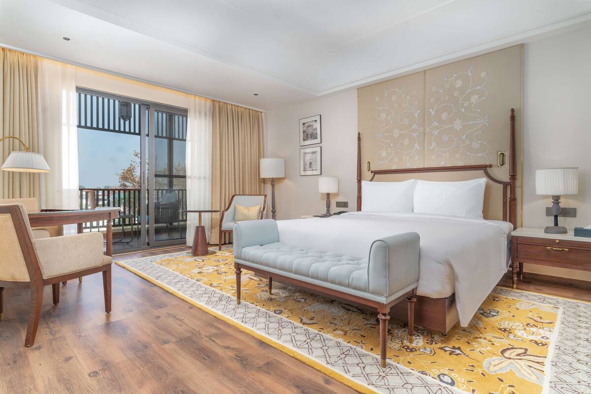 Quarto de hotel com uma cama grande, área de estar com uma cadeira, escrivaninha e varanda com cortinas.