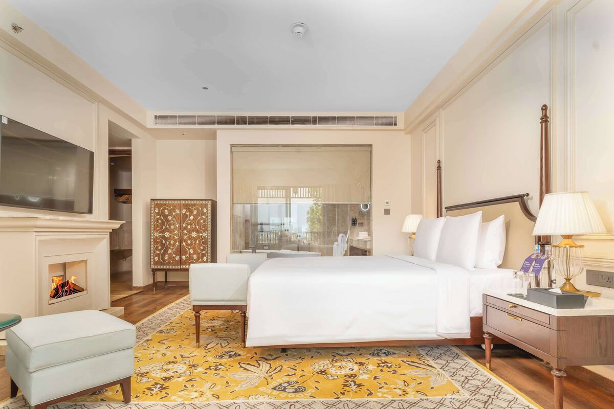 Quarto de hotel com cama, mesa de cabeceira, luminária, televisão e lareira.