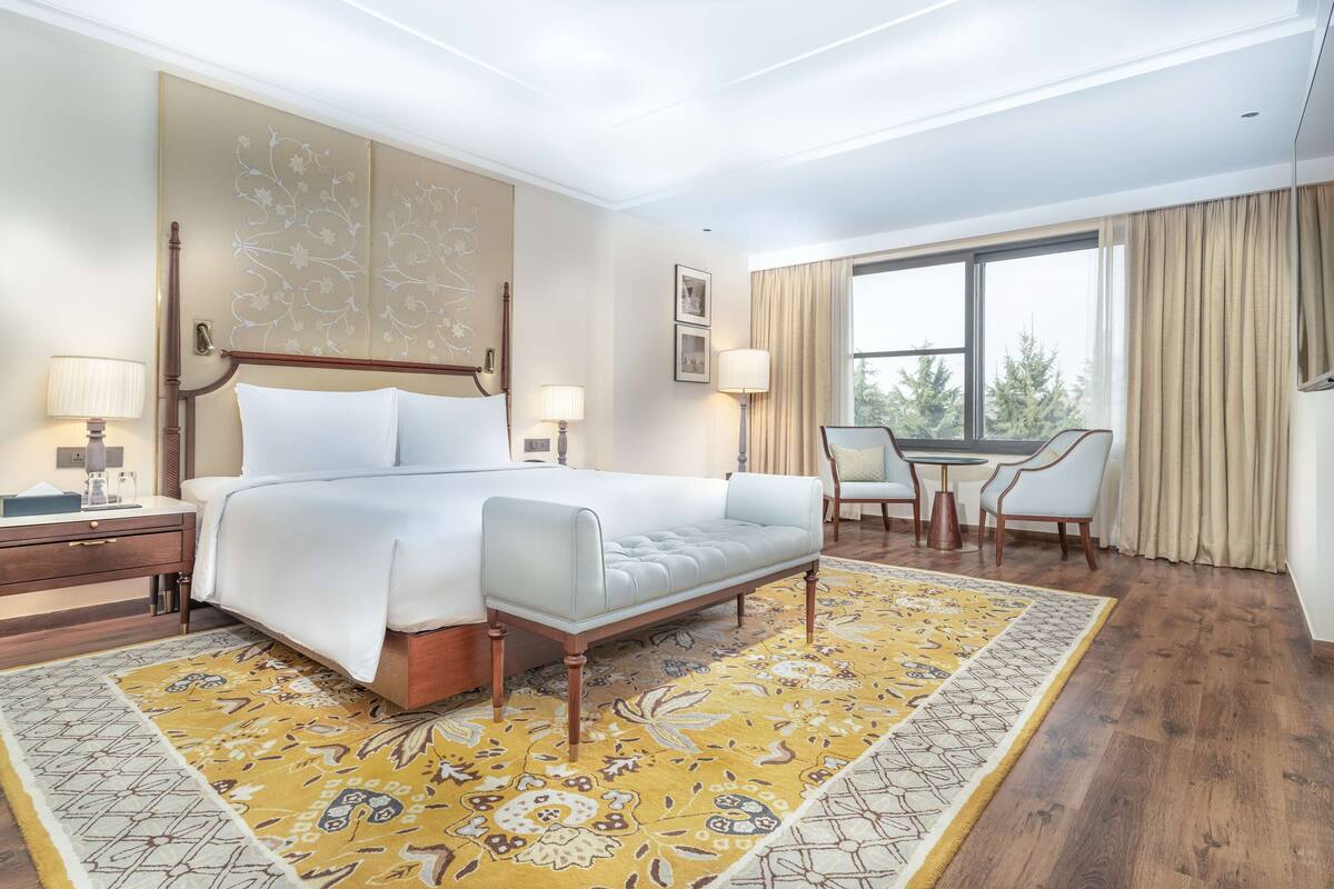 Quarto de hotel com uma cama grande, área de estar, escrivaninha e janela com cortinas.