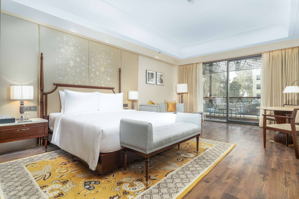 Quarto de hotel com uma cama grande, área de estar, varanda com mesa e cadeiras, e vista para o exterior.