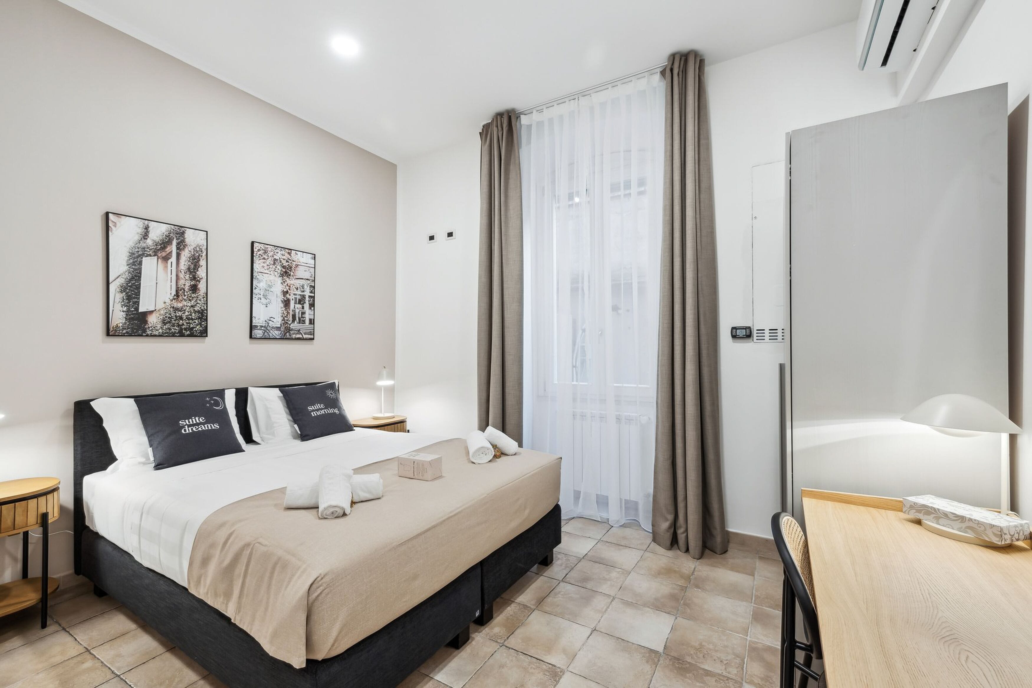 Quarto de hotel moderno com uma cama grande, duas mesas de cabeceira, uma escrivaninha e um espelho amplo.