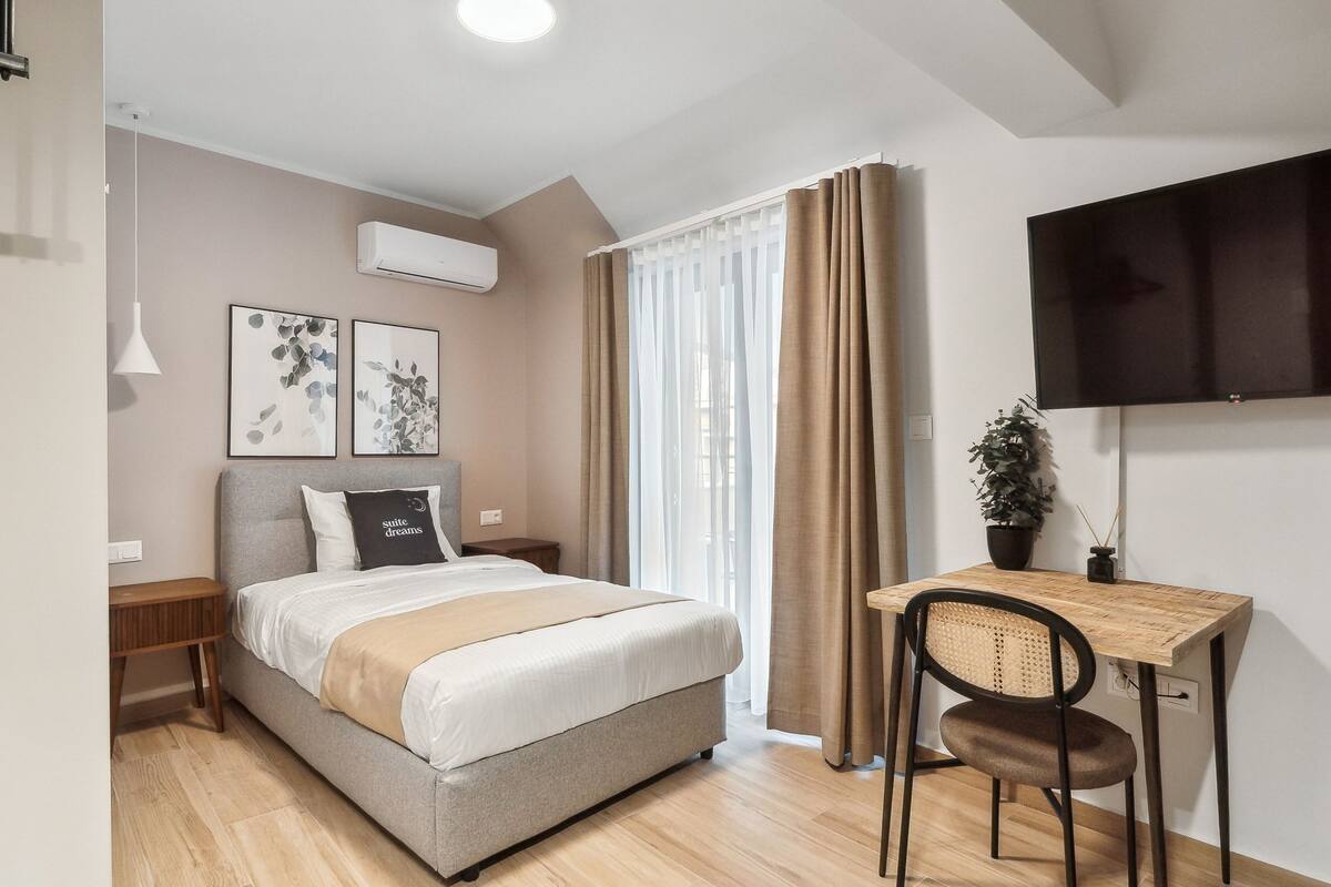 Quarto com cama, mesa de cabeceira, escrivaninha, cadeira, televisão e janela com cortinas.