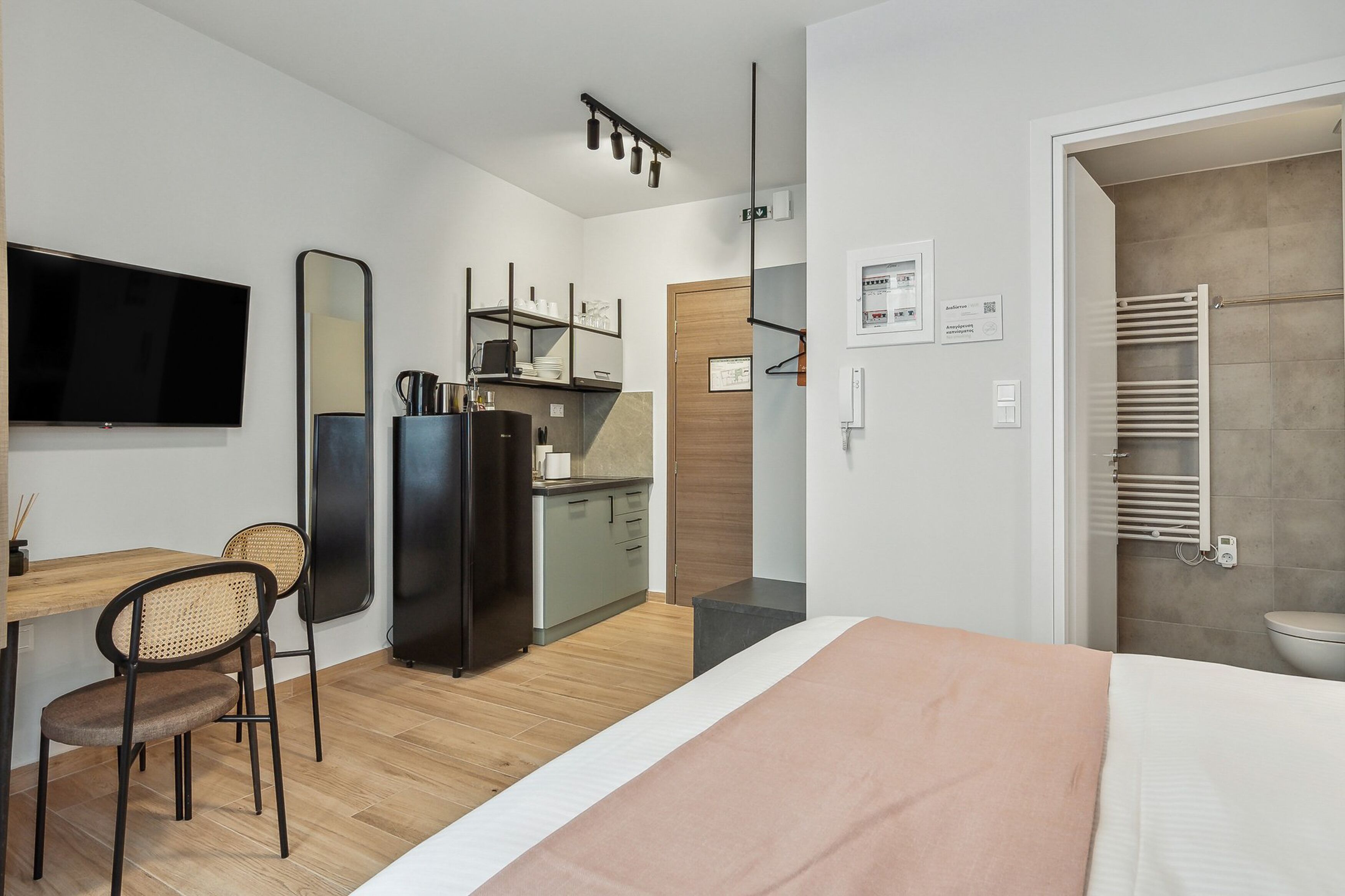 Quarto de hotel moderno com cama, secretária com cadeiras, cozinha compacta e banheiro com porta-toalhas.