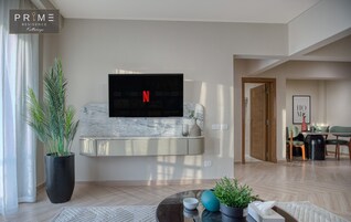 Uma sala de estar moderna com uma TV de tela plana na parede, bancada de mármore, planta em vaso e um espaço de jantar com mesa e cadeiras.