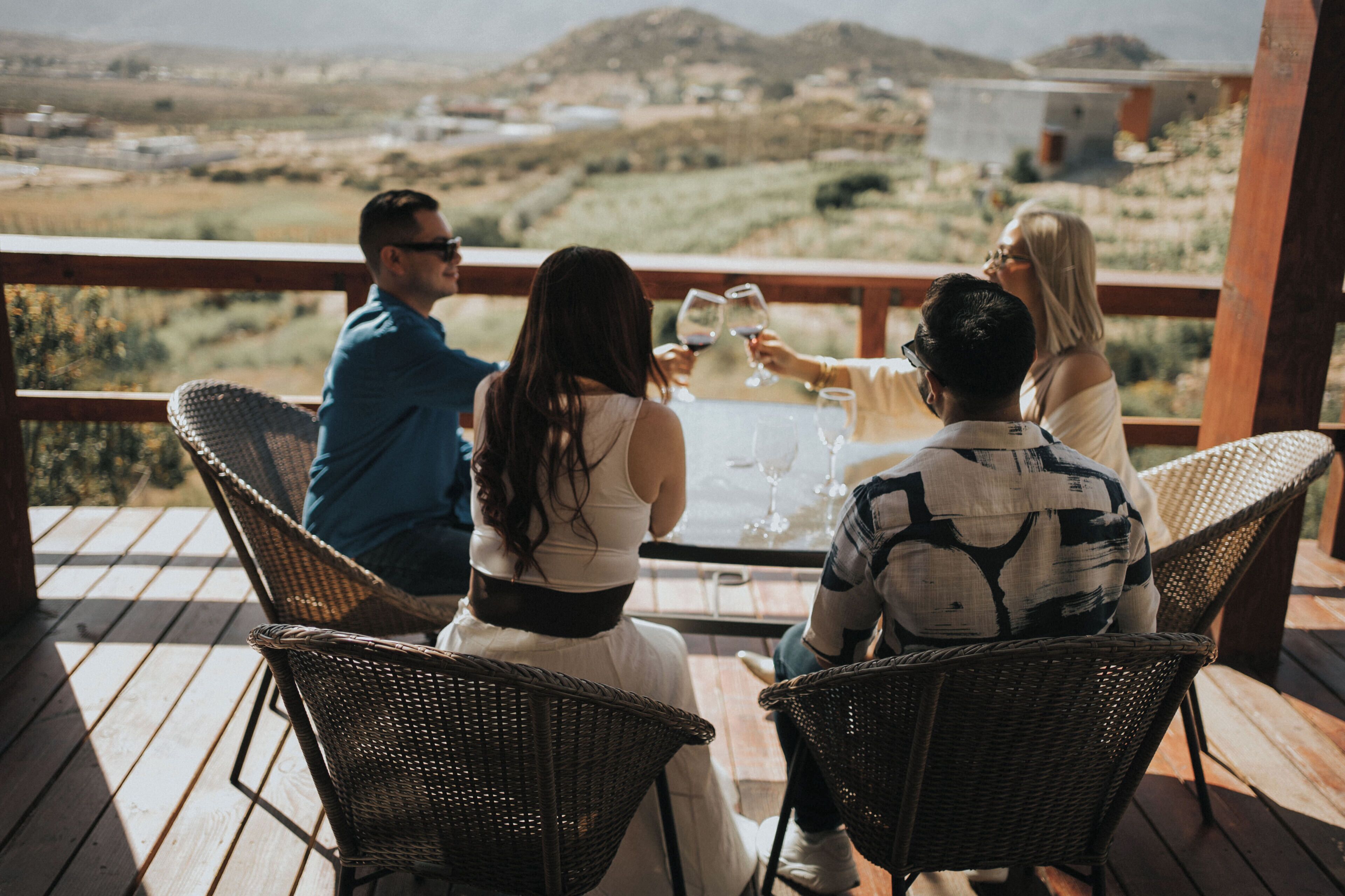Um grupo de pessoas sentado à mesa em um deck de madeira, com cadeiras de vime, com vista para uma paisagem com colinas e edifícios.