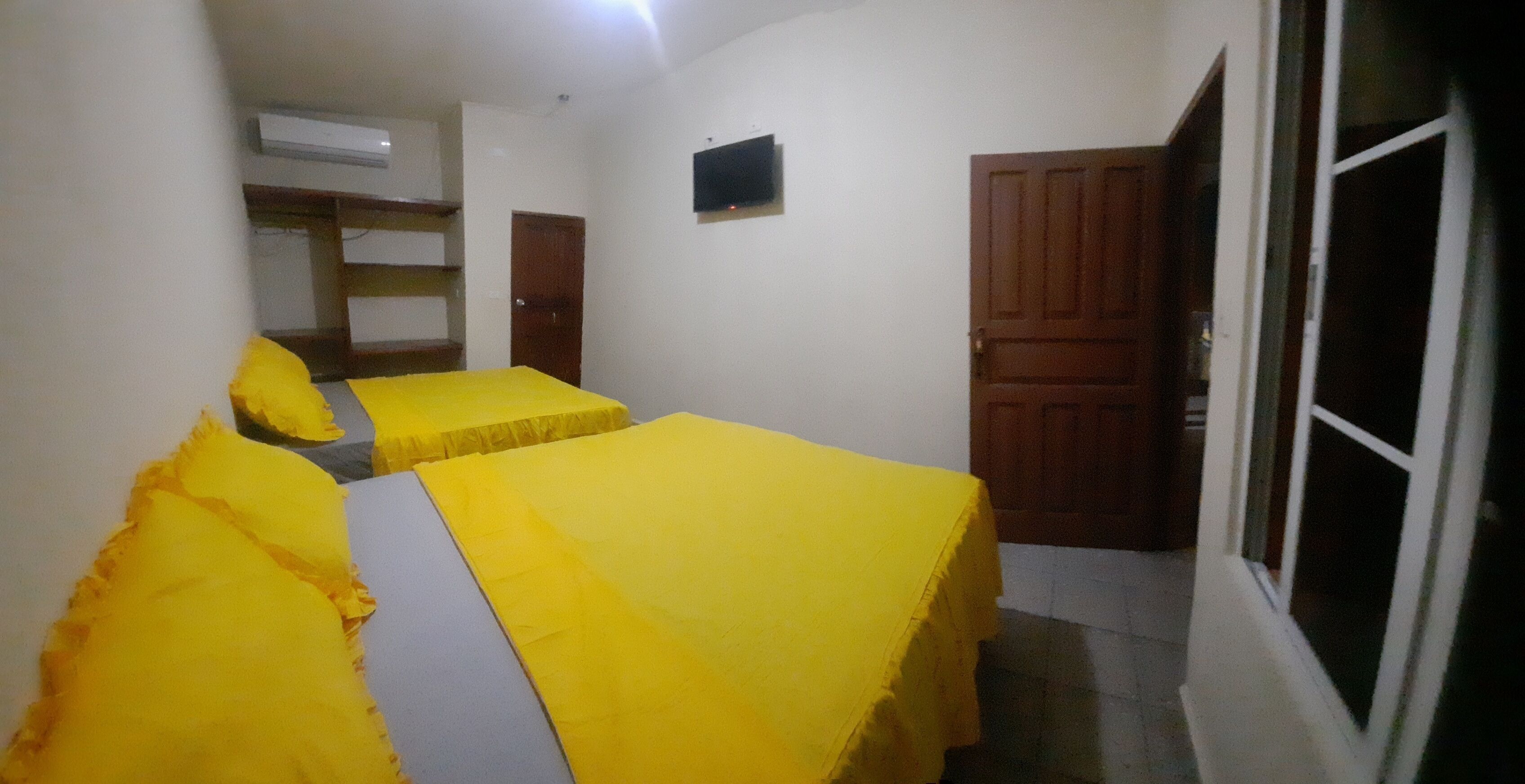 Quarto com duas camas cobertas por lençóis amarelos, uma porta de madeira e uma janela.
