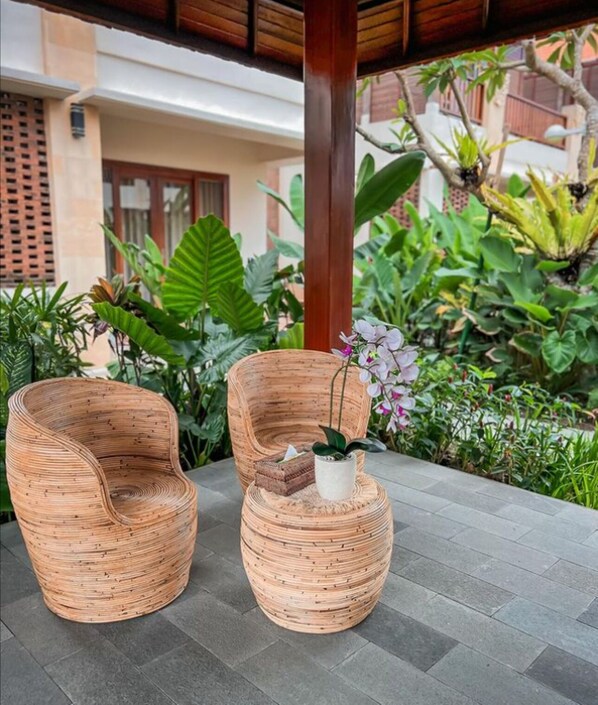 Duas cadeiras de vime com uma pequena mesa e um vaso de planta numa área de jardim com chão de azulejos e estrutura de madeira.