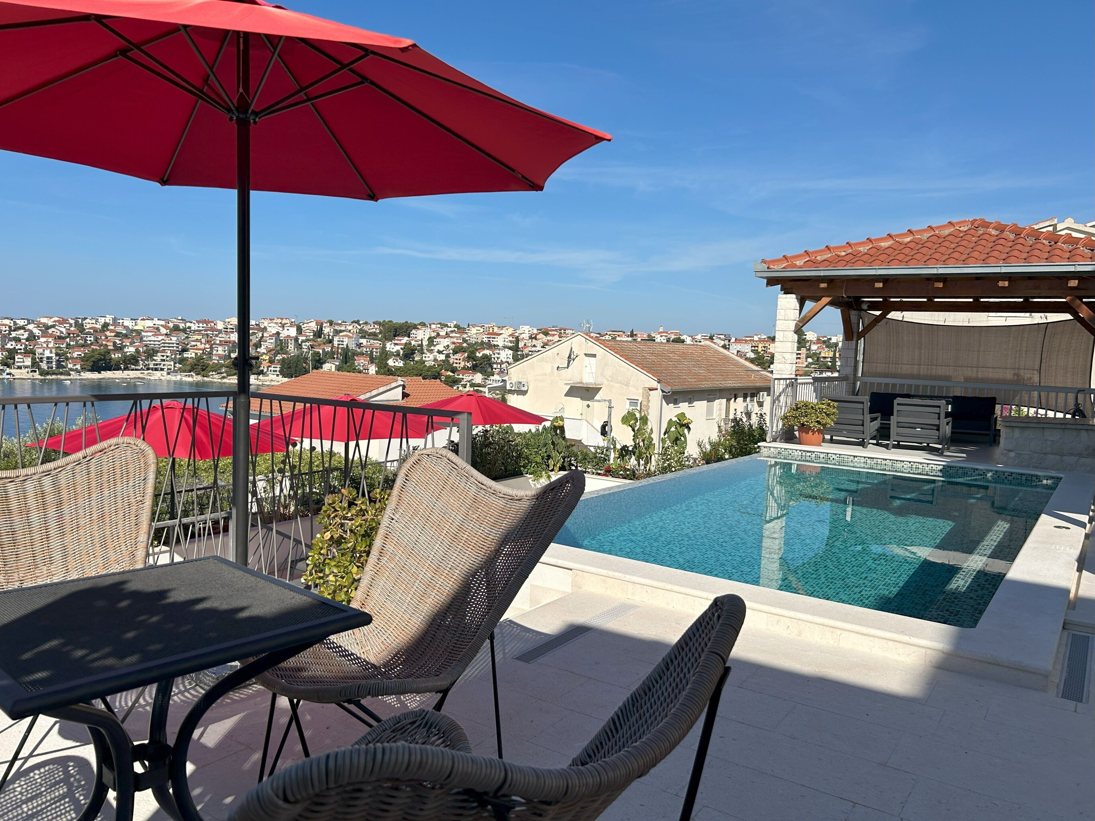 Piscina no terraço com cadeiras de vime e um guarda-sol vermelho, com vista para a cidade.