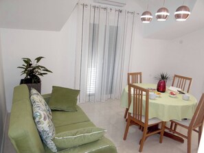 Uma área de refeições com mesa posta para dois, um sofá verde com almofadas estampadas e um vaso de planta.
