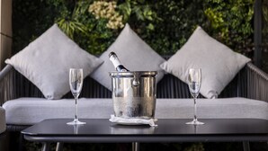 Uma mesa com um balde de metal contendo uma garrafa, dois copos e um guardanapo. A mesa está montada em frente a um banco estofado com almofadas e uma parede verde.