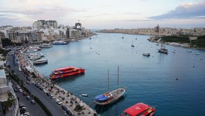 Uma marina com vários barcos, incluindo barcos vermelhos para passeios turísticos, e um horizonte urbano com edifícios e uma estrada à beira-mar.