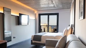 Quarto de hotel moderno com cama, sofá, um espelho grande e varanda com guarda-corpo.
