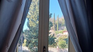 Quarto com uma janela ampla com duas cortinas, uma porta com maçaneta e vista para um jardim ajardinado com árvores e cerca.