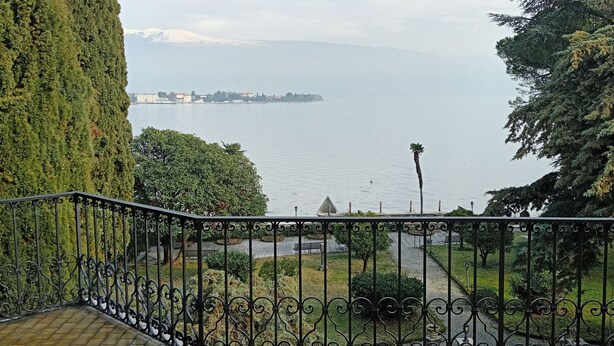 Terraço com vista para um lago e montanhas ao longe, com grades de metal e mobiliário de exterior.