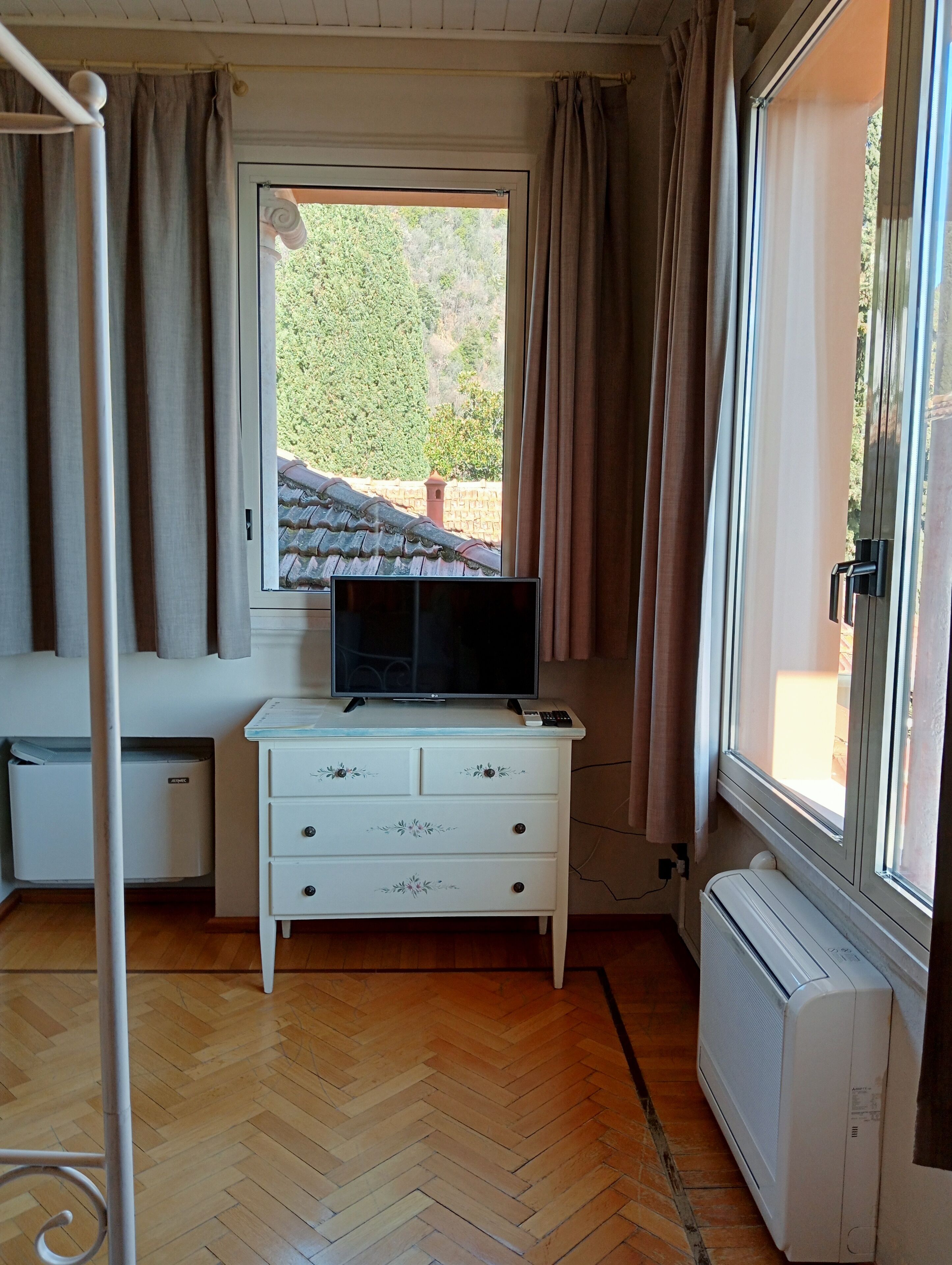 Um quarto com piso de madeira em padrão losango, cômoda branca com detalhes florais, uma televisão em cima e uma janela com cortinas.