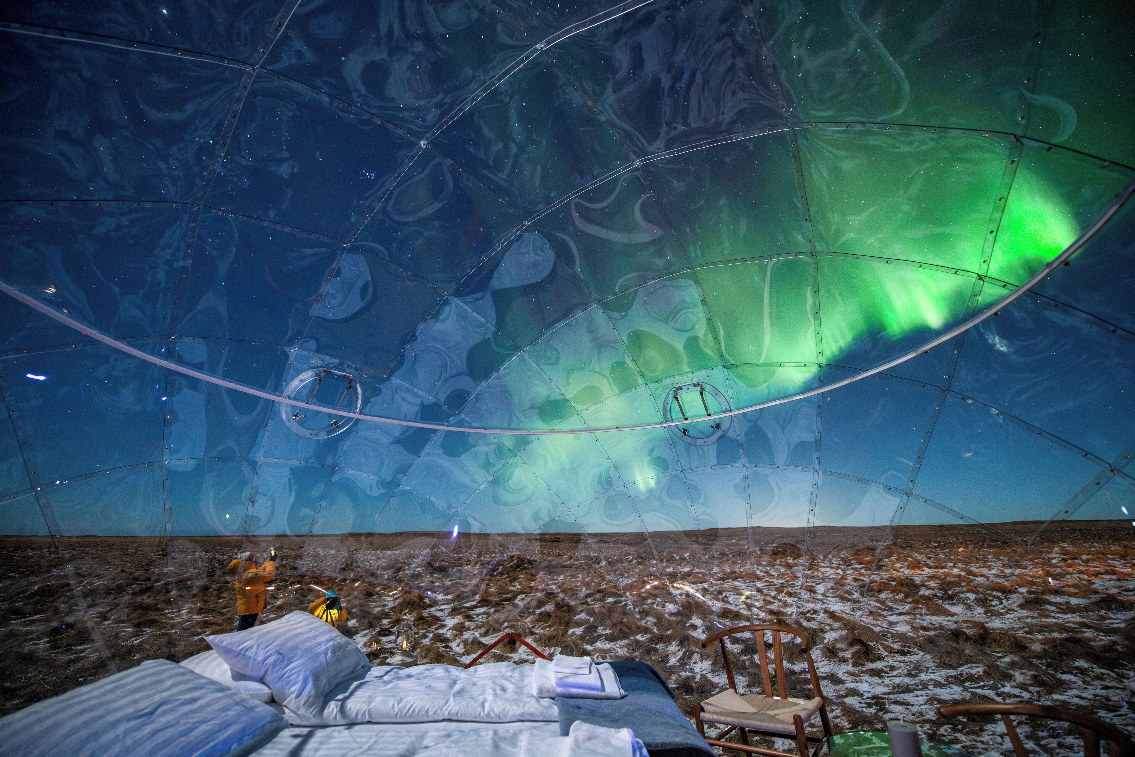 Estrutura em forma de cúpula com uma cama e cadeiras no interior, e vista do céu noturno com luzes verdes.