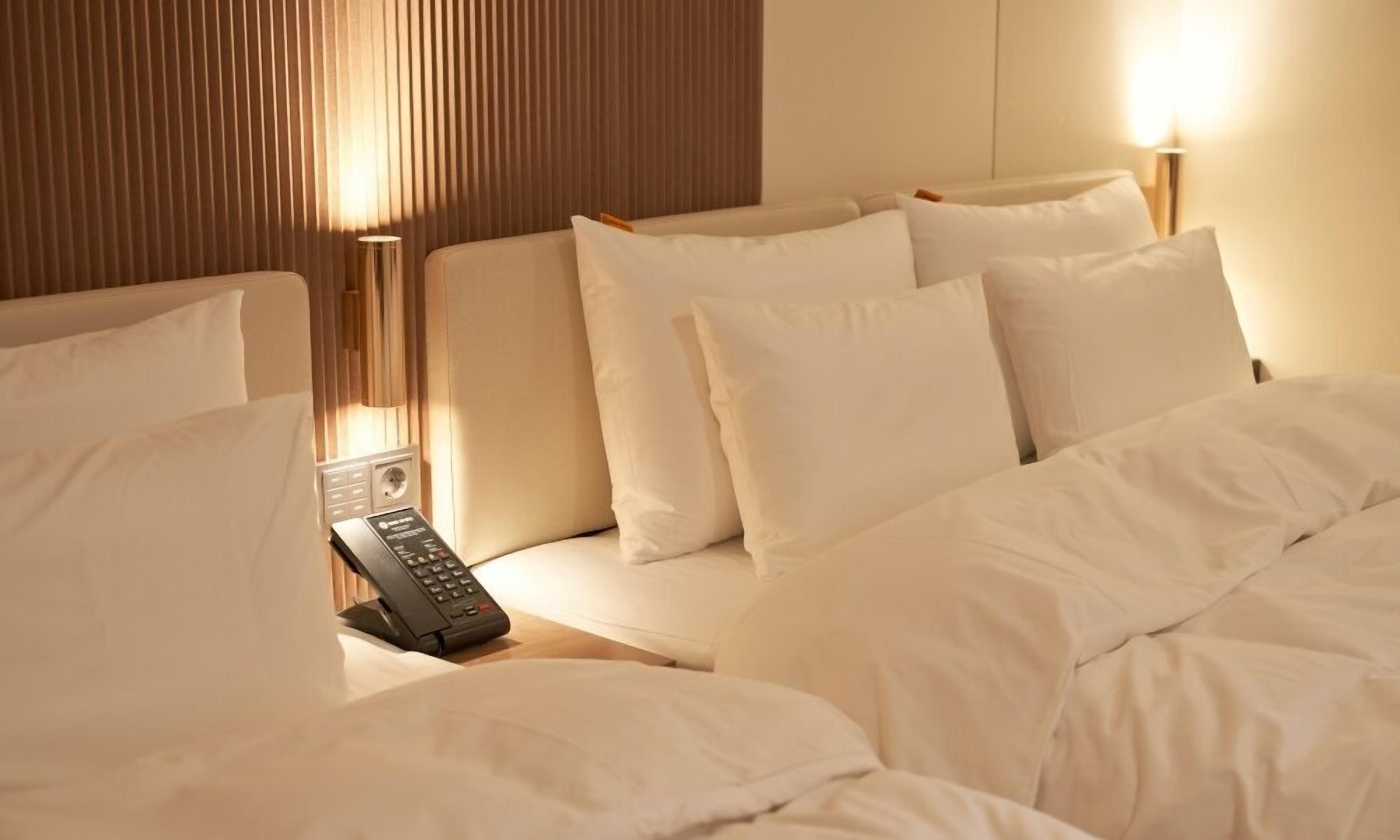 Quarto de hotel com cama, almofadas, comando remoto e luminária de parede.