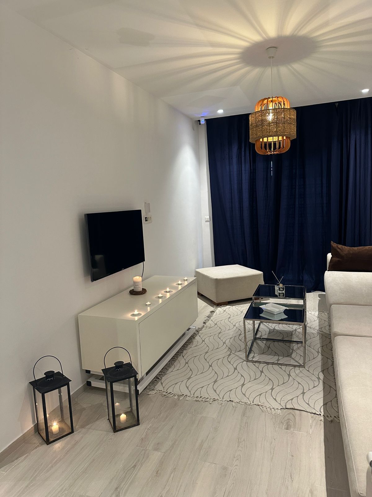 Uma sala de estar moderna com uma TV de tela plana, um console branco com velas, uma mesa de centro de vidro e uma cortina azul escura.
