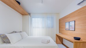 Quarto de hotel moderno com cama bem arrumada, escrivaninha de madeira e janela grande com cortinas leves.