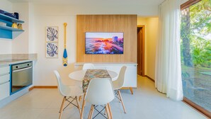 Cozinha moderna com forno embutido, área de refeições com mesa e cadeiras, TV montada e vista para o verde através de uma janela grande.