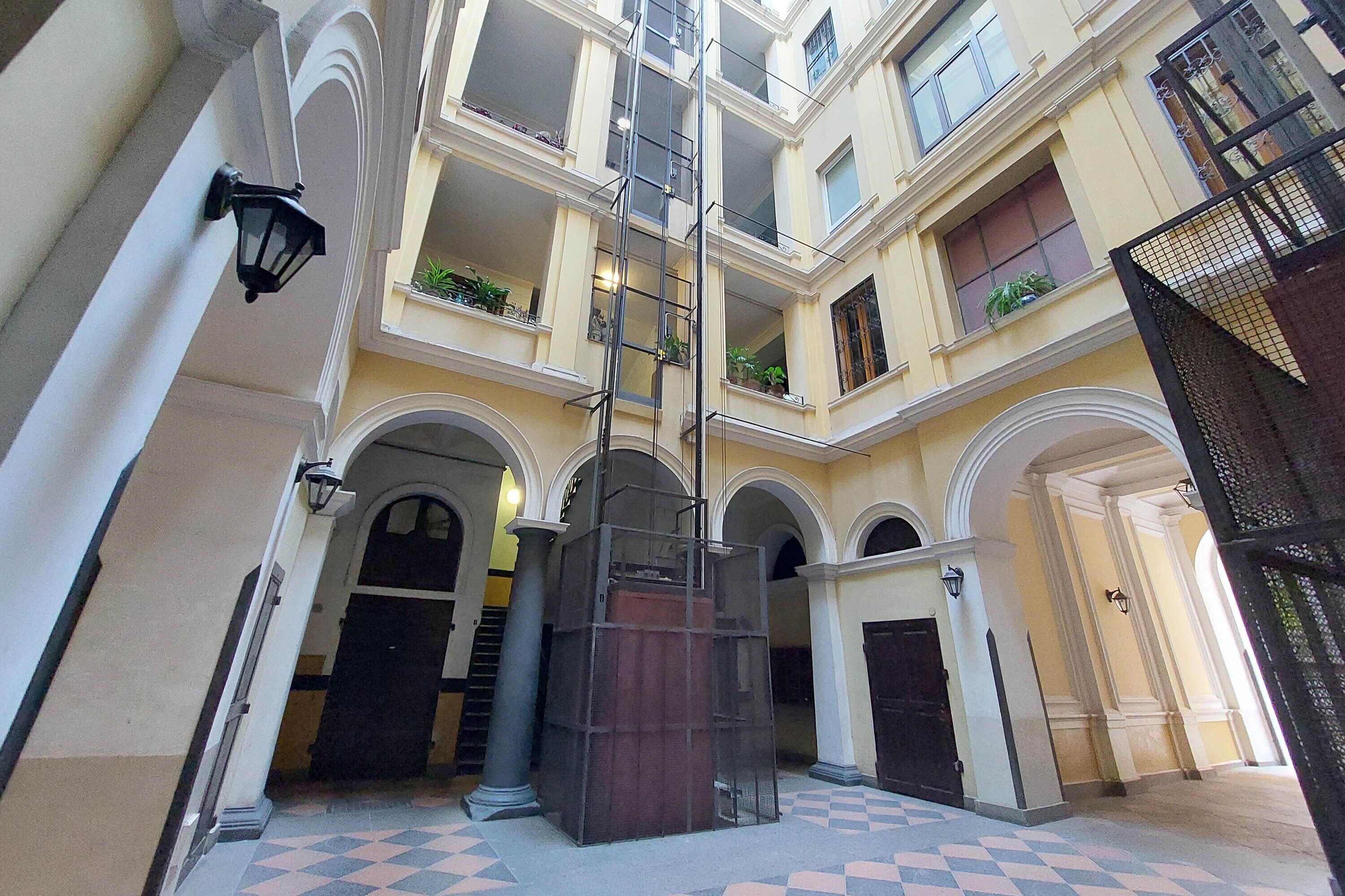 Um átrio com vários níveis, com escadaria central, varandas com plantas e um padrão de piso xadrez.