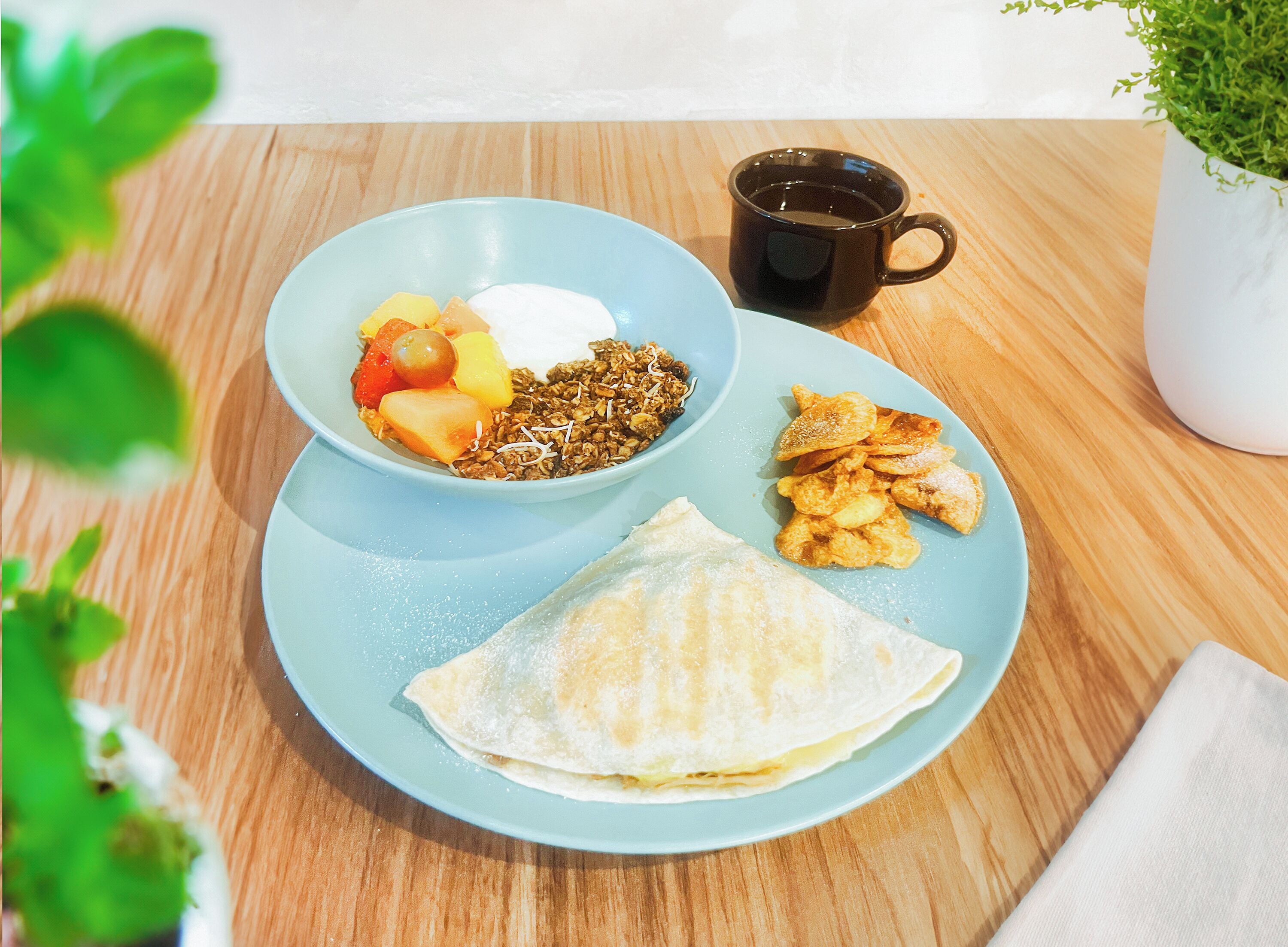Prato de pequeno-almoço com uma crepe, fruta, iogurte, granola e uma chávena de café sobre uma mesa de madeira.