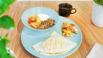 Prato de pequeno-almoço com uma crepe, fruta, iogurte, granola e uma chávena de café sobre uma mesa de madeira.