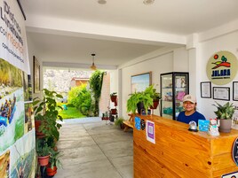 Balcão de recepção de hotel com bancada de madeira, uma pessoa atrás do balcão e vista para um jardim com plantas em vasos e um muro de tijolos.