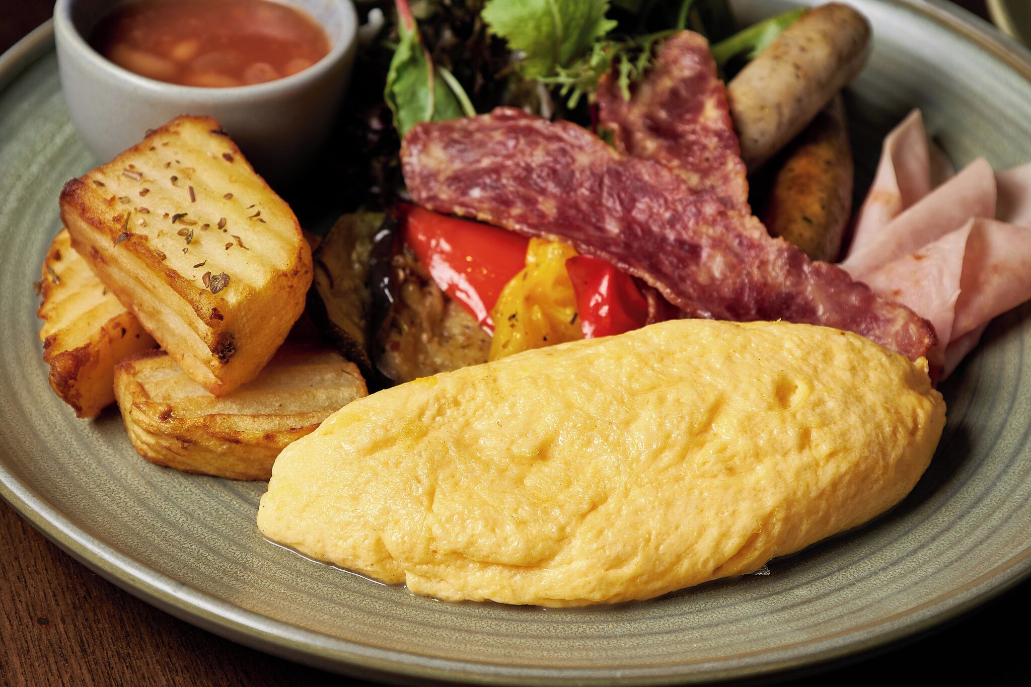 Um prato com ovos mexidos, batatas assadas, uma fatia de fiambre, um pedaço de carne vermelha e uma pequena tigela de molho.