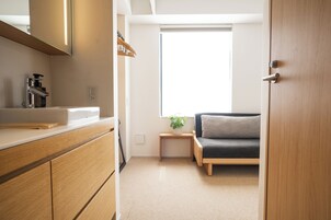 Quarto moderno e compacto com um armário de madeira, uma pequena mesa de cabeceira com uma planta e um sofá cinza.
