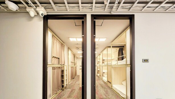 Corredor de hotel com portas espelhadas, carpete estampado e teto com tubulações aparentes.