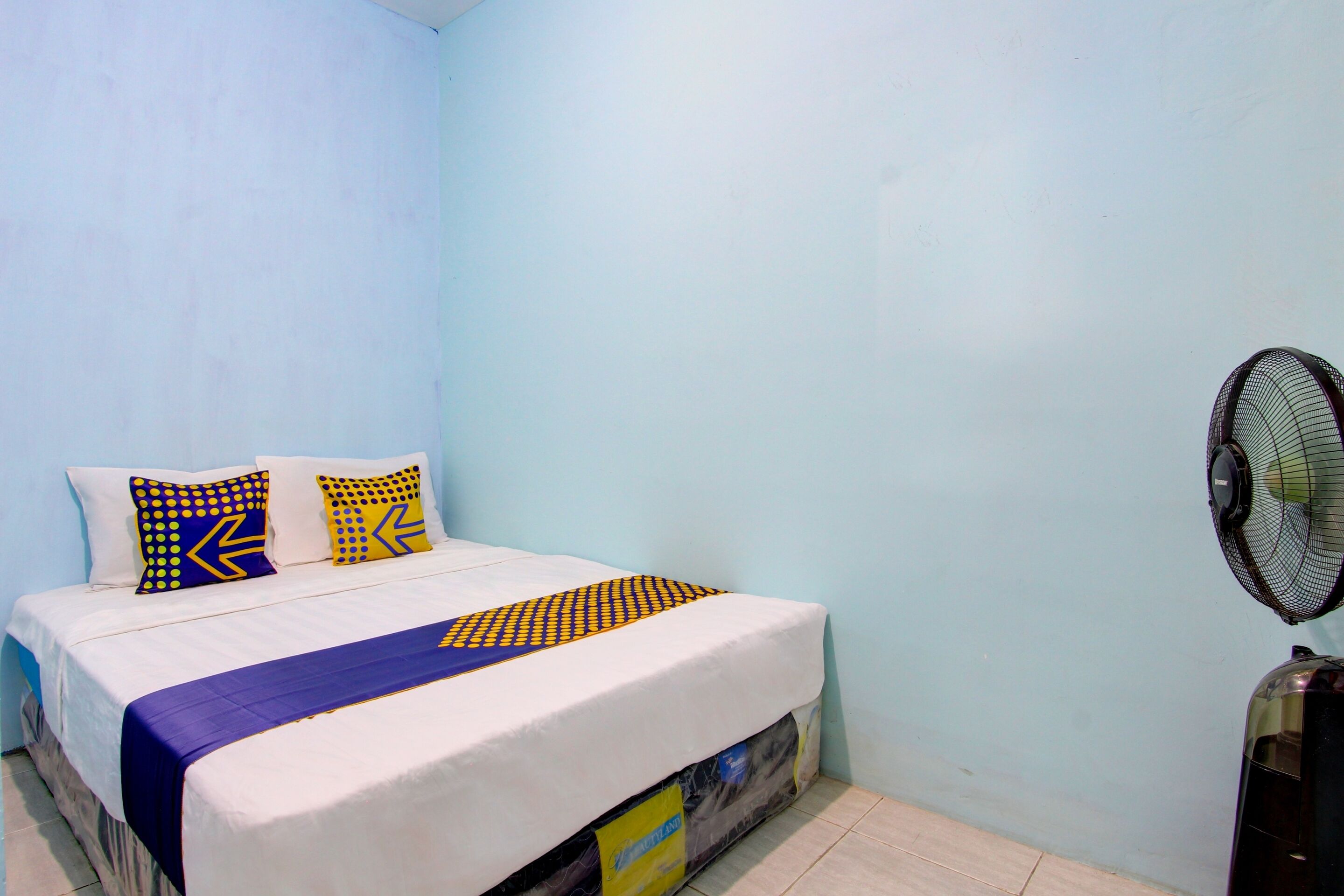 Uma cama com colcha branca e azul, duas almofadas com estampas amarelas, e um ventilador na parede.