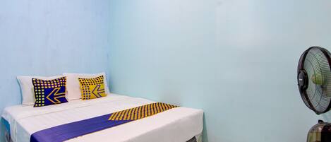 Uma cama com colcha branca e azul, duas almofadas com estampas amarelas, e um ventilador na parede.