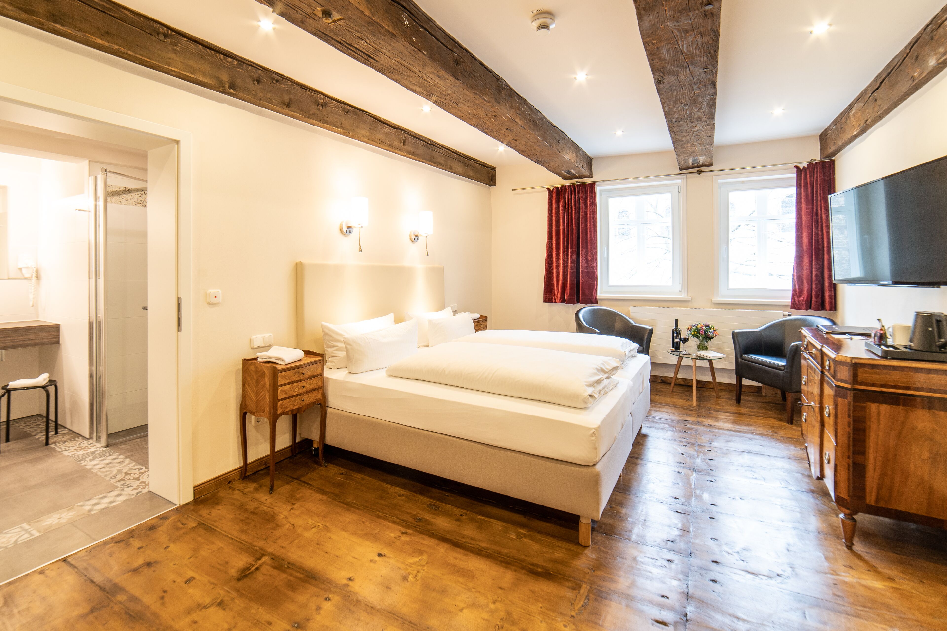 Quarto de hotel com piso de madeira, uma cama grande, uma escrivaninha com cadeira, uma televisão e uma janela com cortinas.