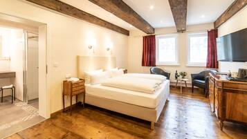 Quarto de hotel com piso de madeira, uma cama grande, uma escrivaninha com cadeira, uma televisão e uma janela com cortinas.