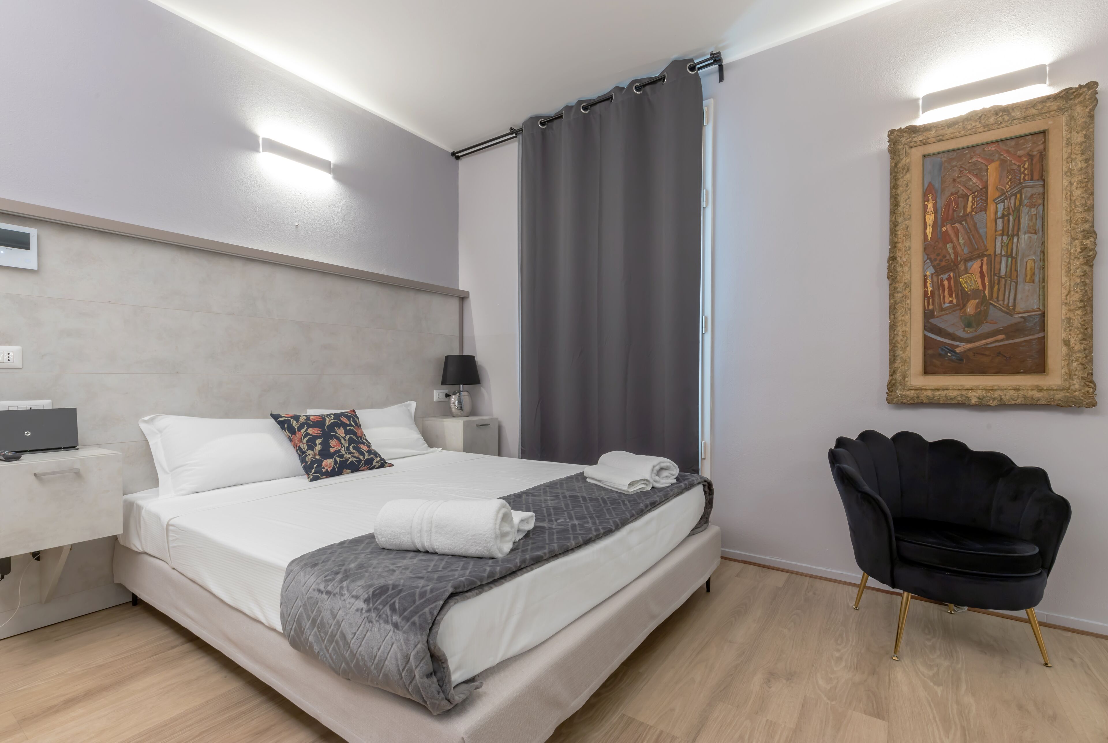 Quarto de hotel moderno com uma cama bem arrumada, uma cadeira preta e um quadro na parede.